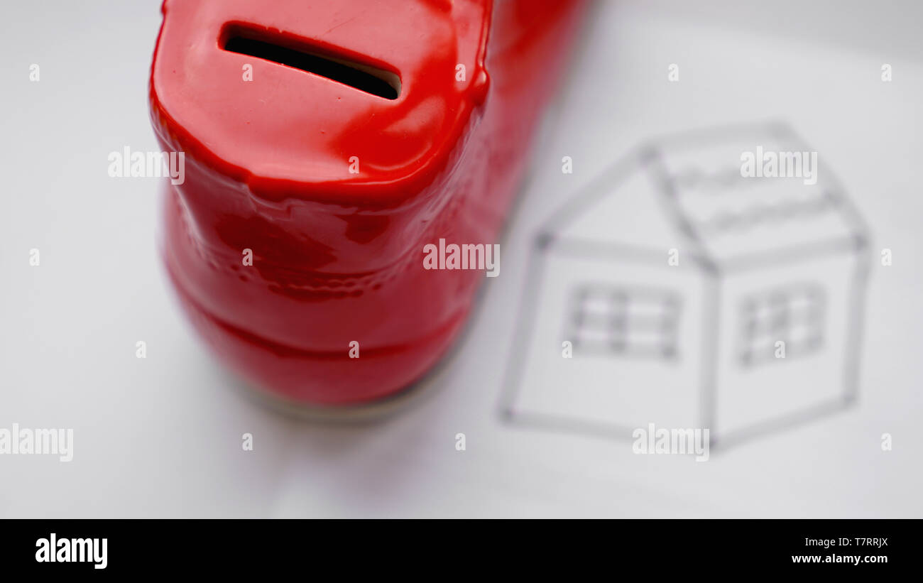 Hand dropping coin into the moneybox. Drawing a house on a white sheet ...