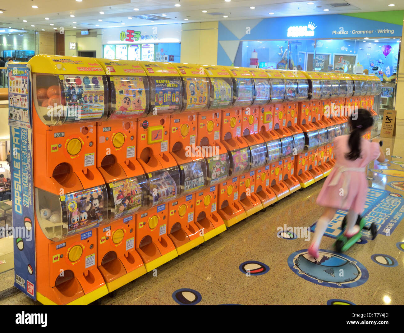 Small girl rides a scooter to run pass an array of capsule vending ...