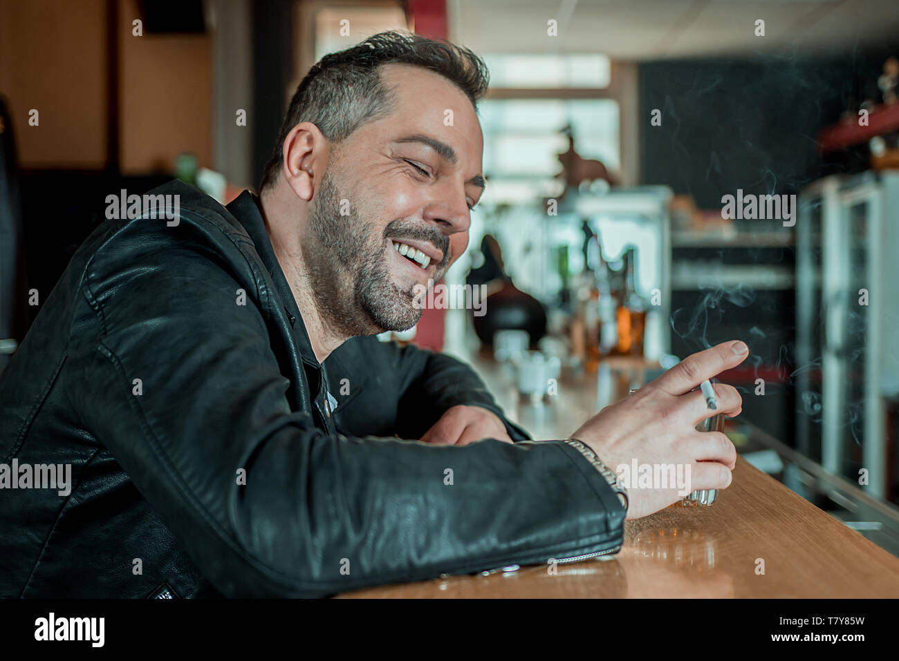 A laughing man in a bar at the counter with a whiskey and a cigarette ...