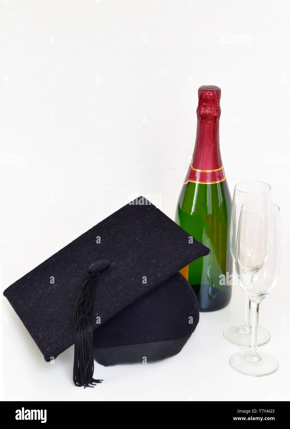 Graduation cap, champagne bottle and glasses on table with a white wall ...