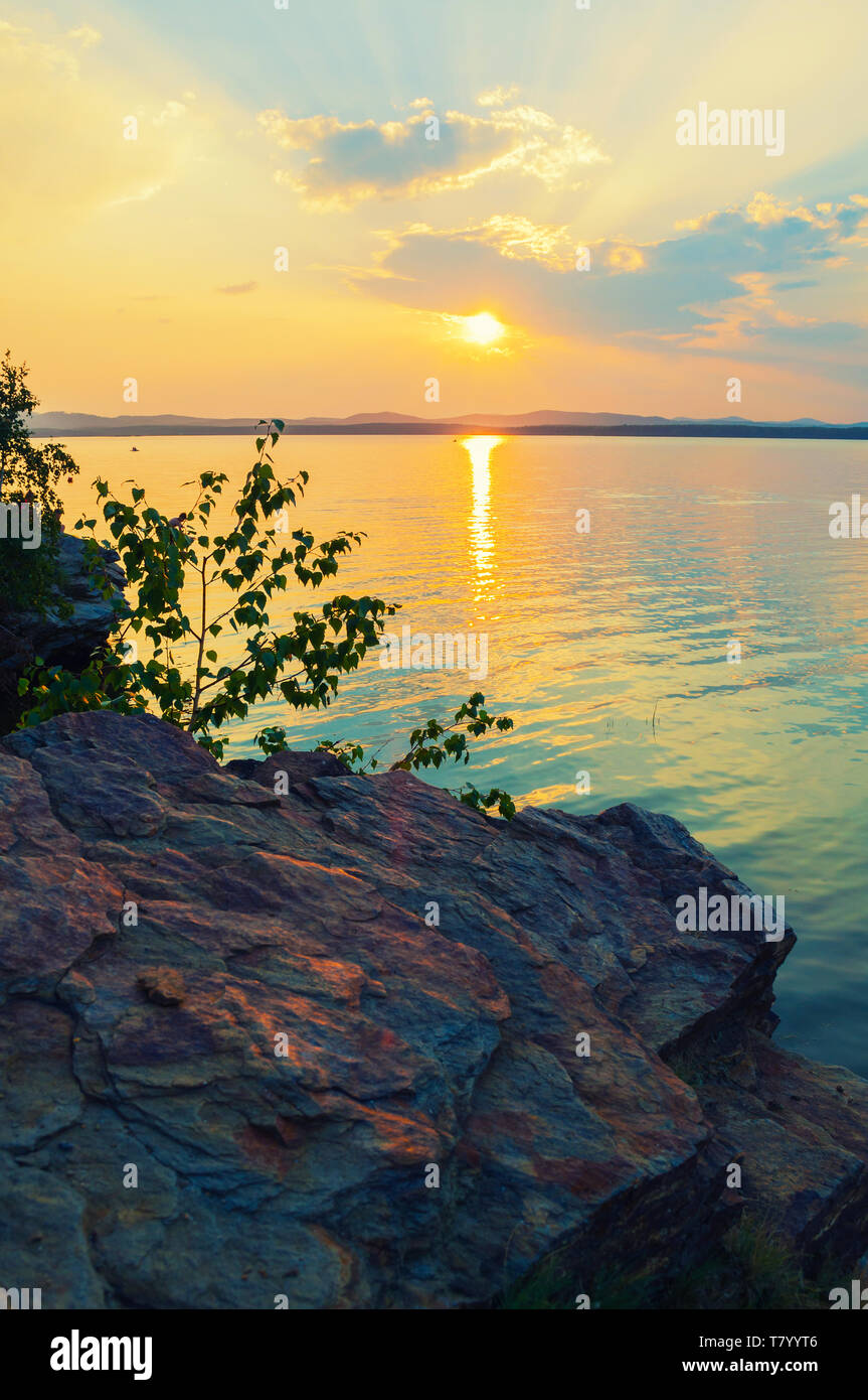 Summer sunset landscape - stone cliff and lake lit by gold sunset light ...