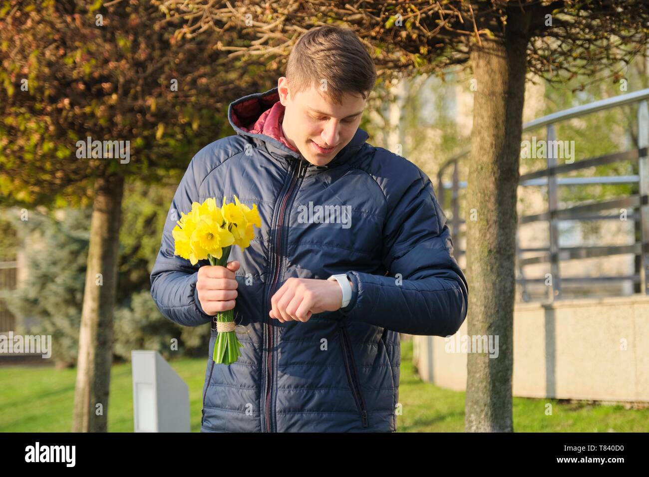 Young man with bouquet of spring flowers looks at wrist watch, urban ...
