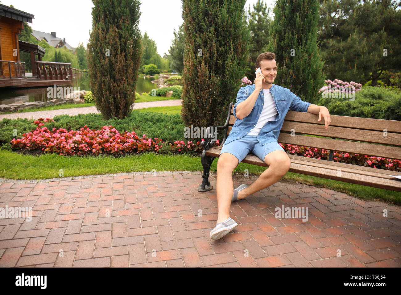 Young man talking by mobile phone while sitting on wooden bench in park ...