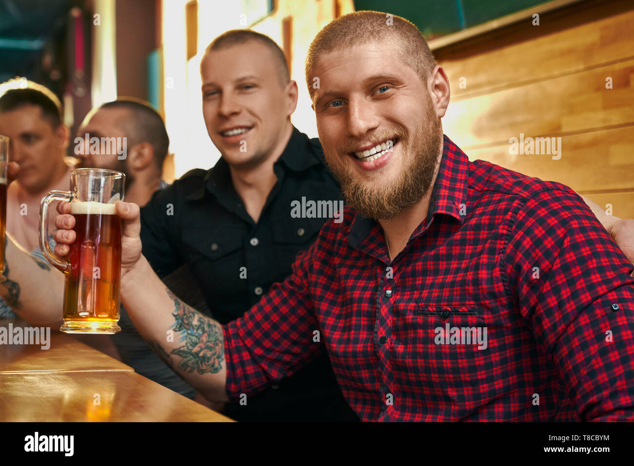 Happy male hipster with tattoo keeping glass of beer, looking at camera ...
