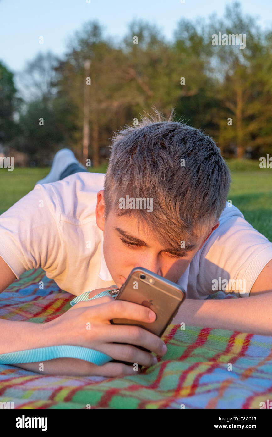 A teenage boy looks at his mobile phone whilst lying on a rug outside ...