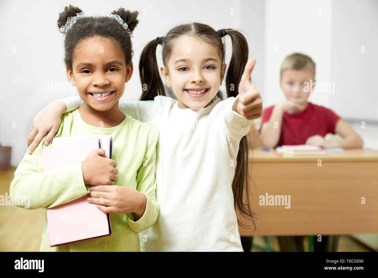 Pretty schoolgirls standing in classroom, classmates showing thumbs up ...