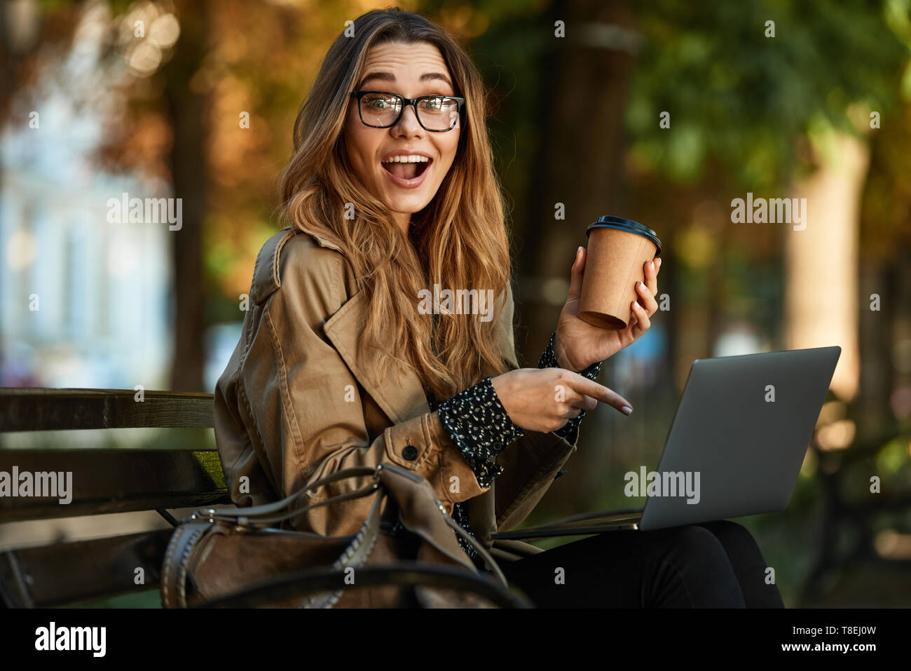 Photo of excited woman 20s wearing coat and eyeglasses working on ...
