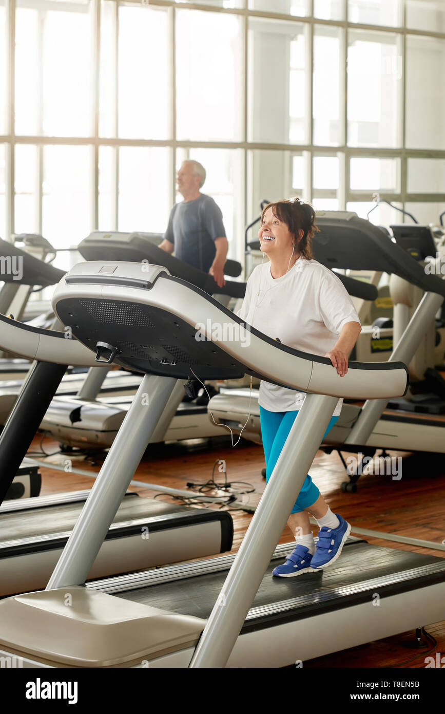 Smiling senior woman running on treadmill in gym. Cheerful aged woman ...