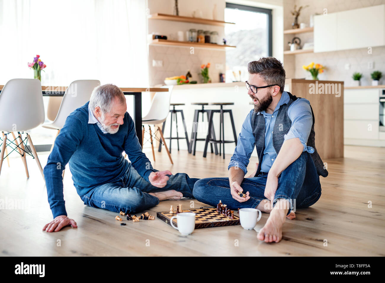 An Adult Hipster Son And Senior Father Sitting On Floor Indoors At Home