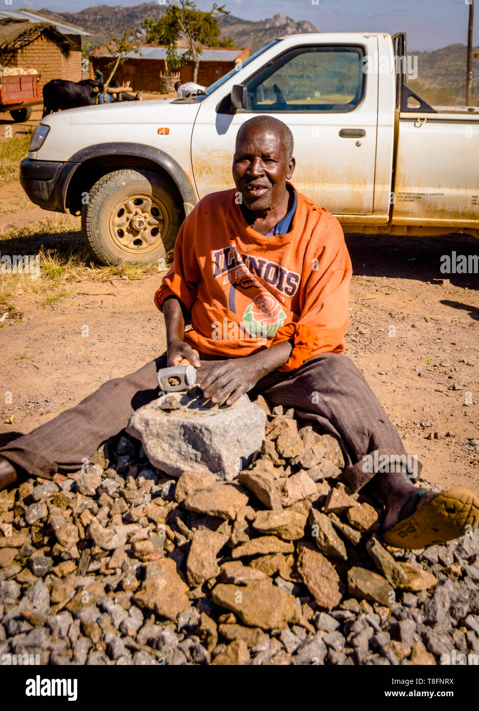 old man sits on the ground breaking rocks for sale with a hammer in hot ...