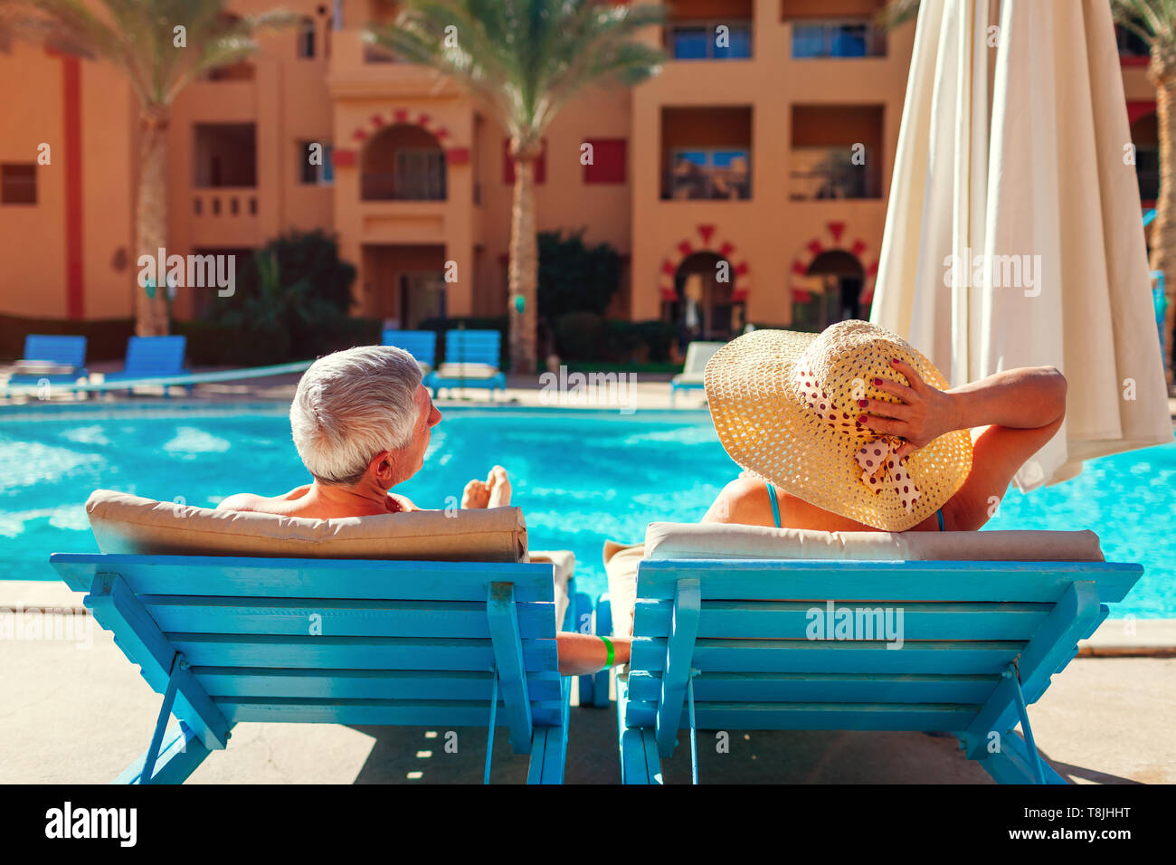 Senior Couple Relaxing By Swimming Pool Lying On Chaise Longues People