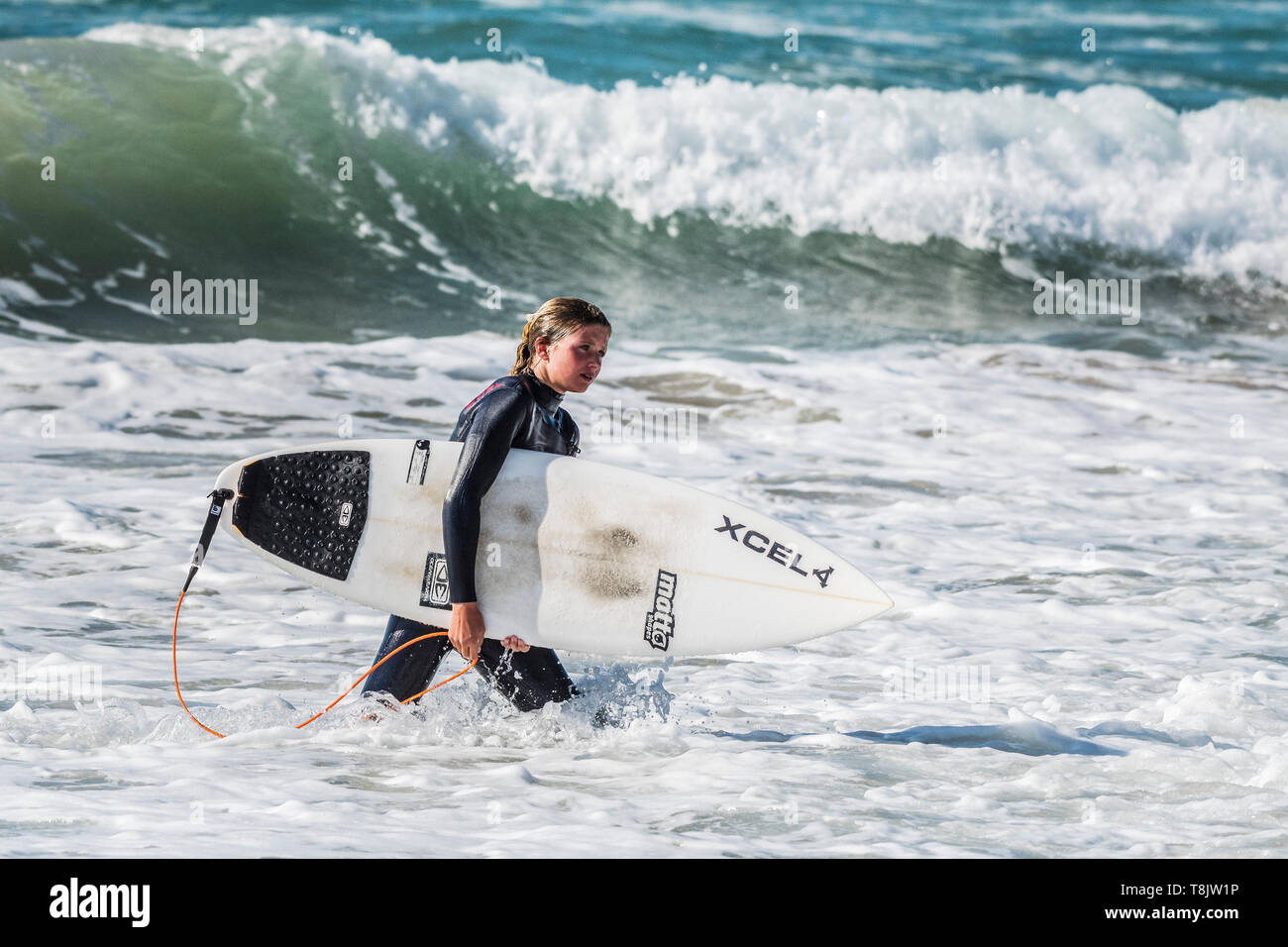 A young tired female surfer carrying her surfboard and walking out of ...