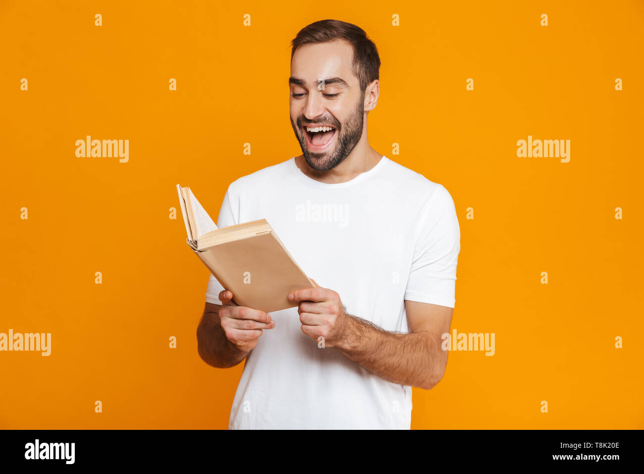 Image of happy man 30s in white t-shirt holding and reading book ...