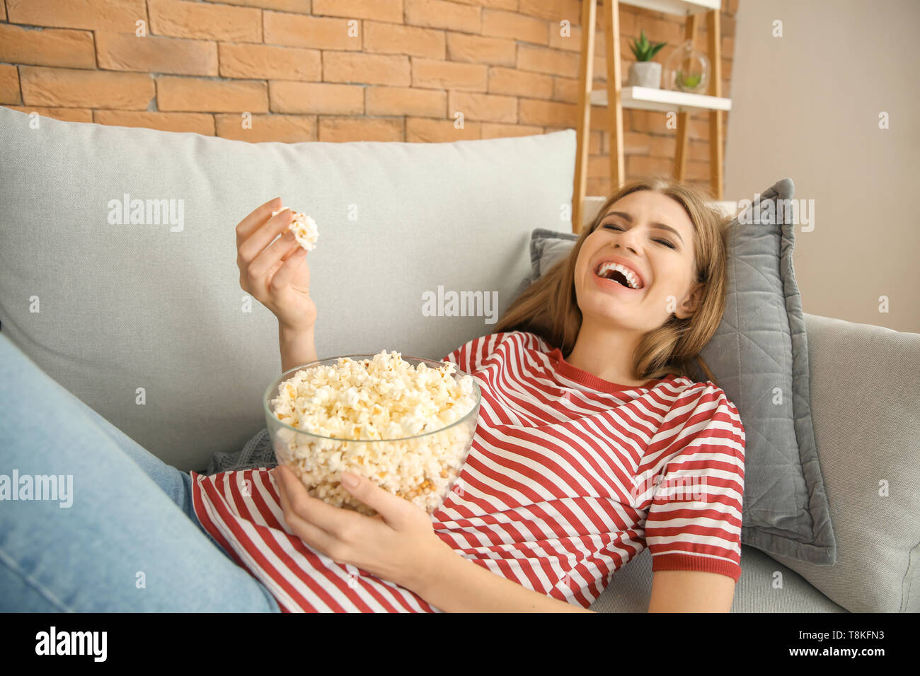 Emotional young woman eating popcorn while watching TV at home Stock ...