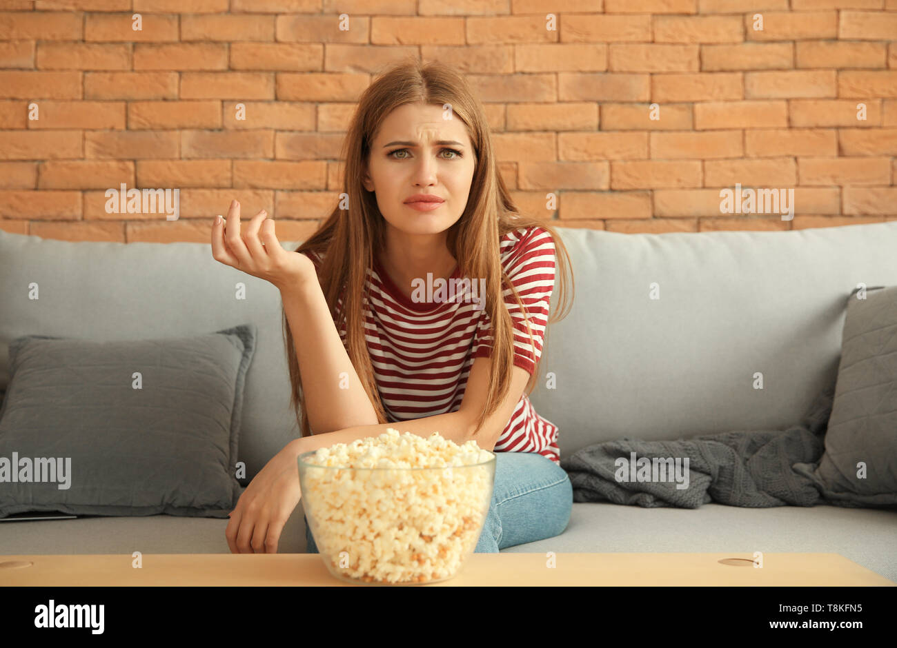 Emotional young woman eating popcorn while watching TV at home Stock ...