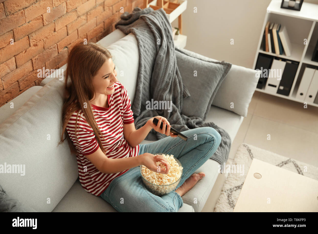 Emotional young woman eating popcorn while watching TV at home Stock ...