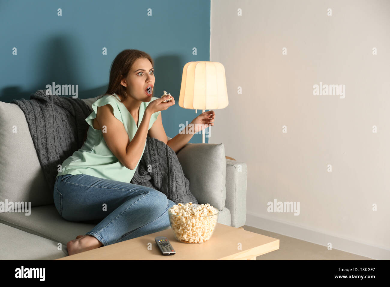 Emotional young woman eating popcorn while watching TV at home Stock ...