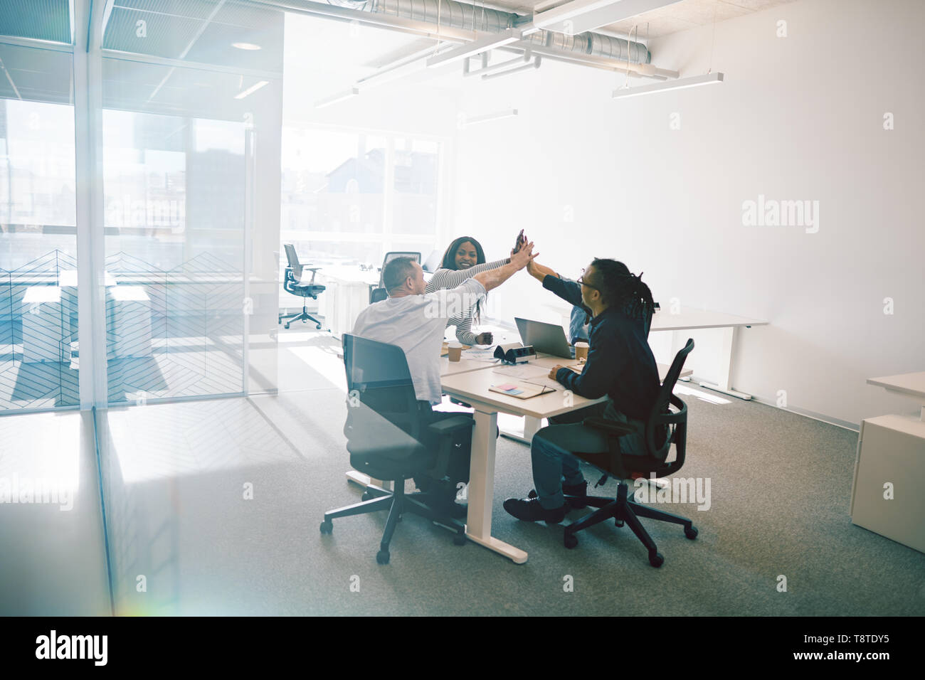Diverse group of work colleagues laughing and high fiving together while sitting around a table ...