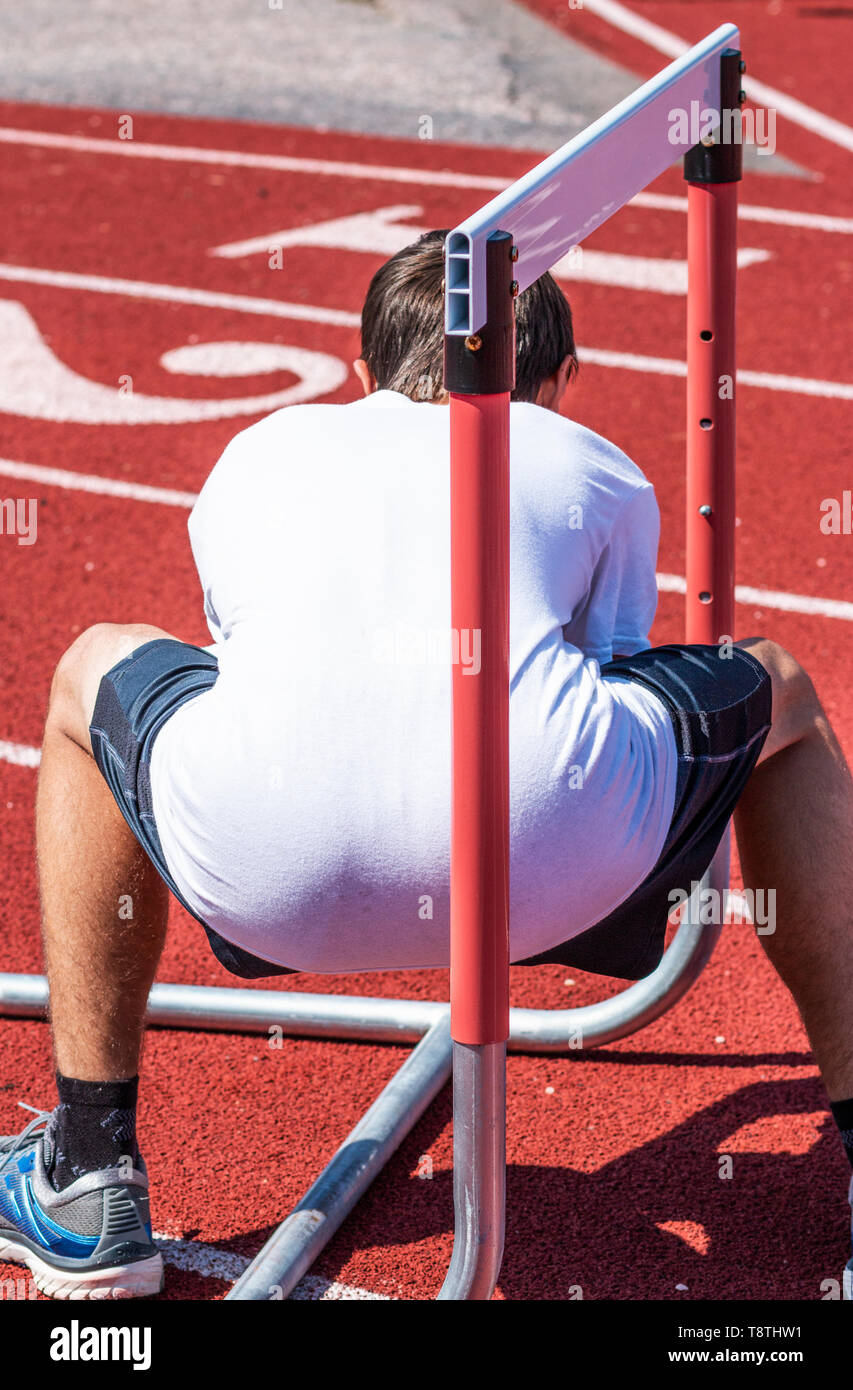 A young athlete is squatting underneath a track hurdle during strength ...