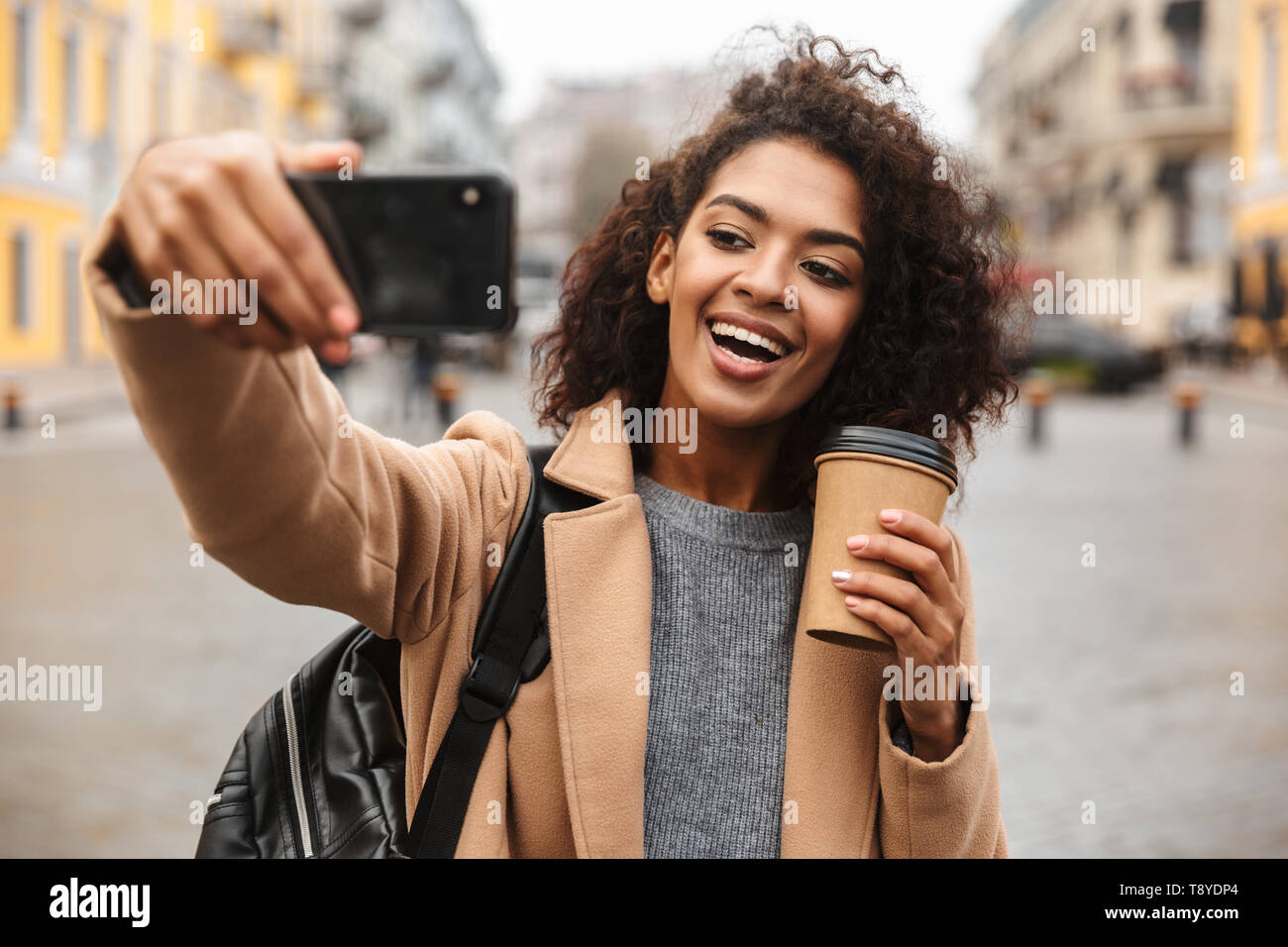 Cheerful young african woman wearing coat walking outdoors, holding takeaway coffee cup, taking ...