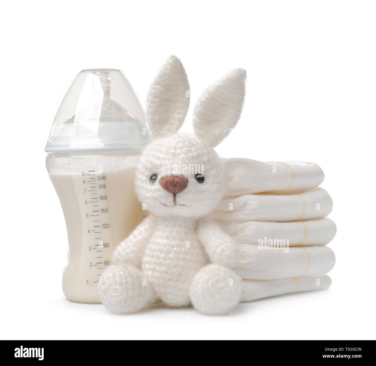Baby bottle with milk, bunny toy and stack of diapers on white ...