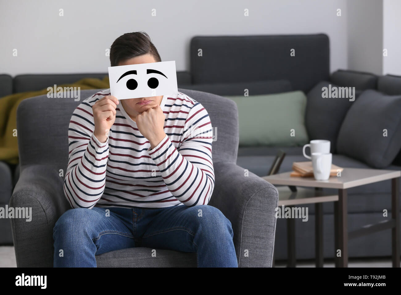 Emotional young man hiding face behind sheet of paper with drawn eyes ...