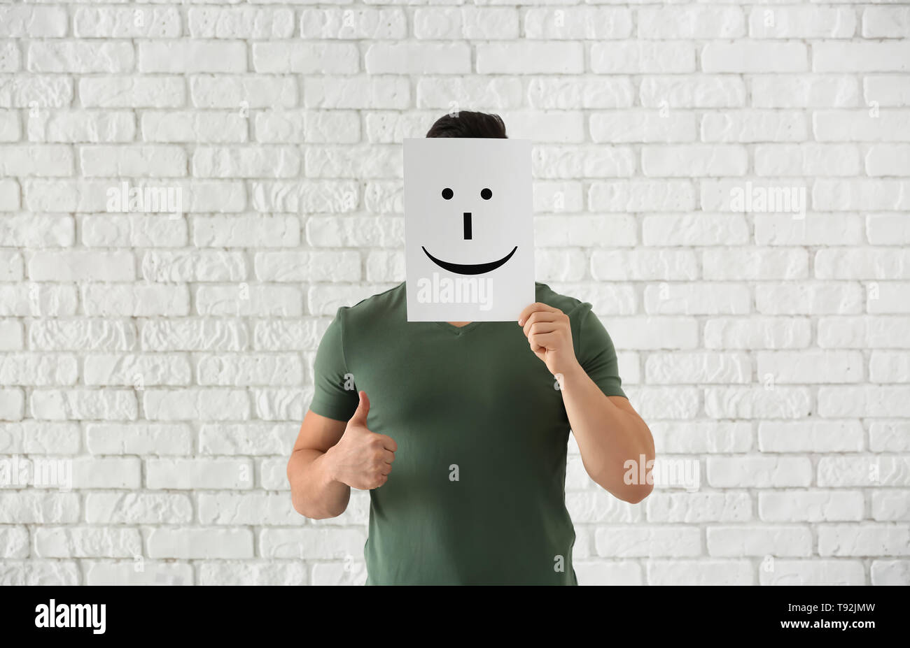 Young man hiding face behind sheet of paper with drawn emoticon against ...