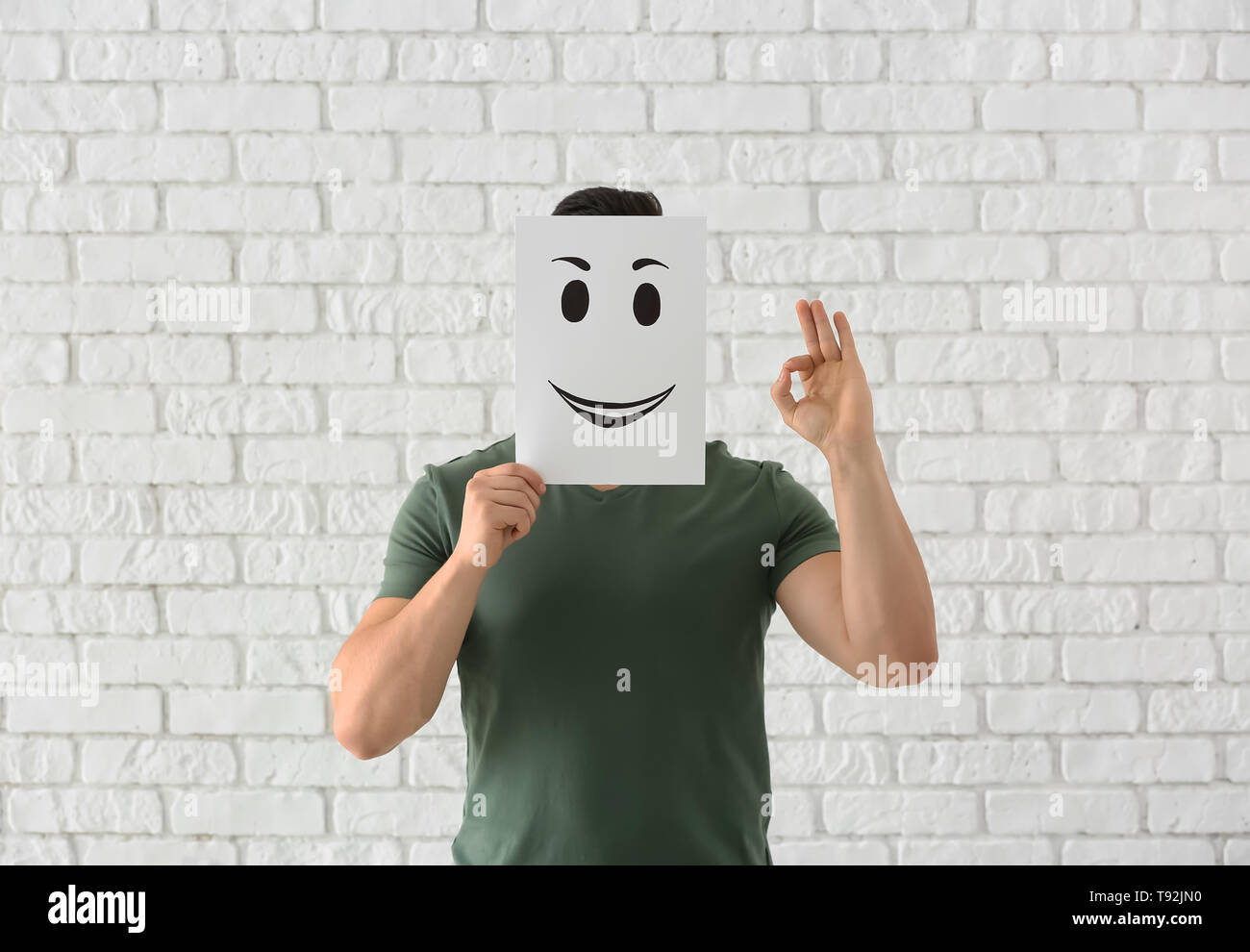 Young man hiding face behind sheet of paper with drawn emoticon against ...