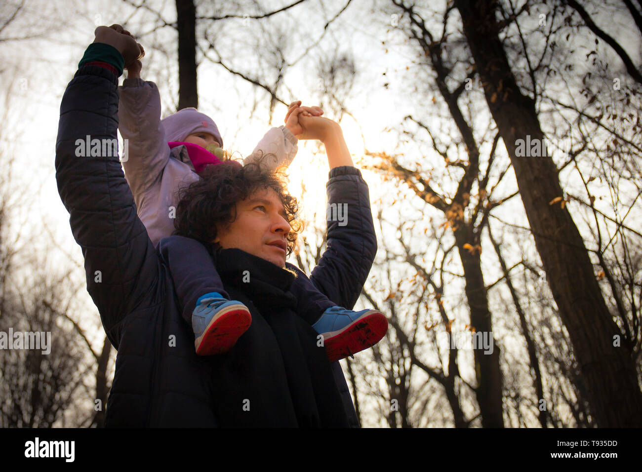 Young dad carrying baby on his shoulders in a forest holding her hands ...