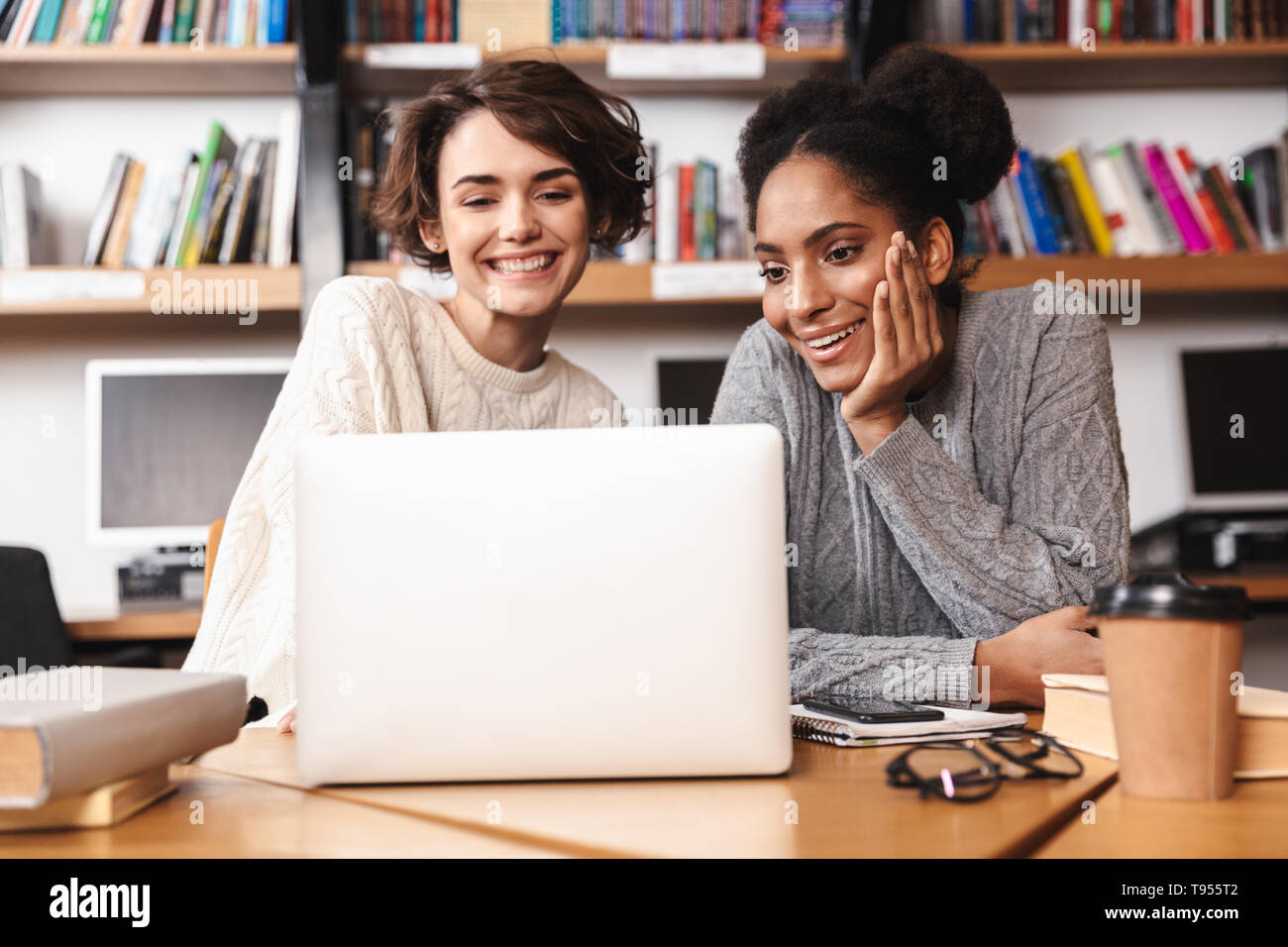 Two cheerful young girls students studying at library, sitting at the ...
