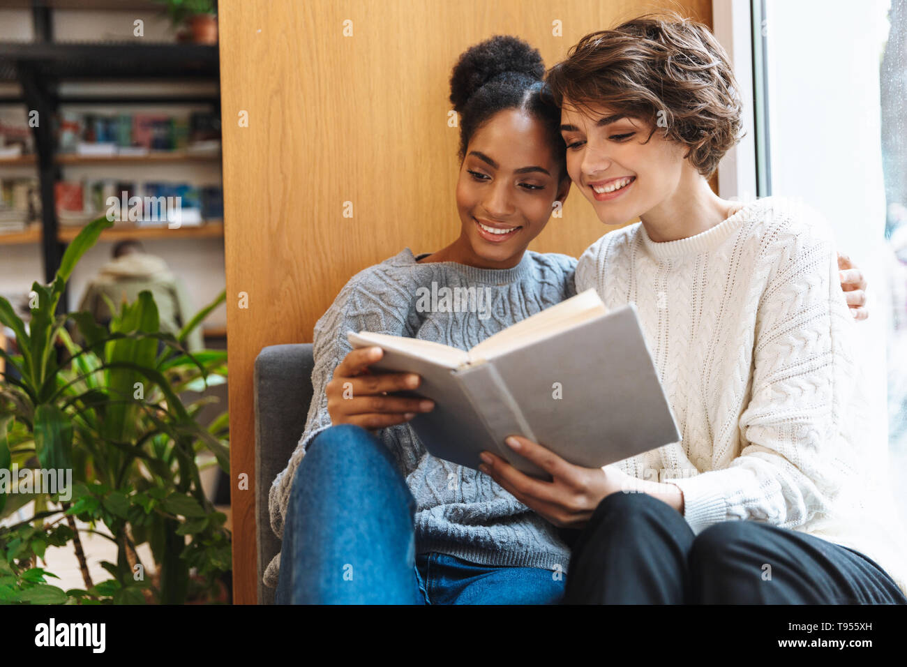 Two cheerful young girls students studying at library, reading a book ...