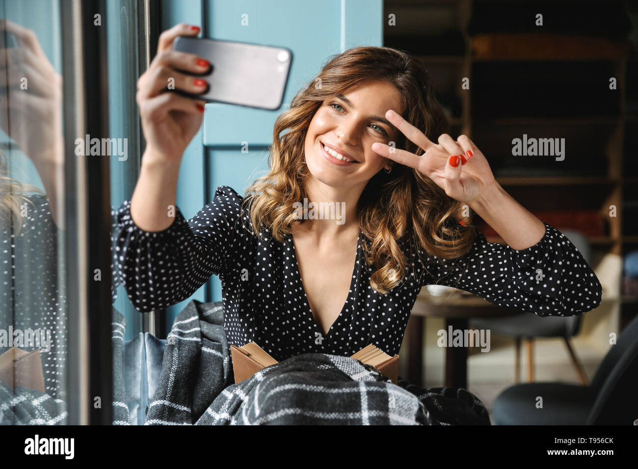 Smiling young woman sitting wrapped in blanket at the window, taking a ...