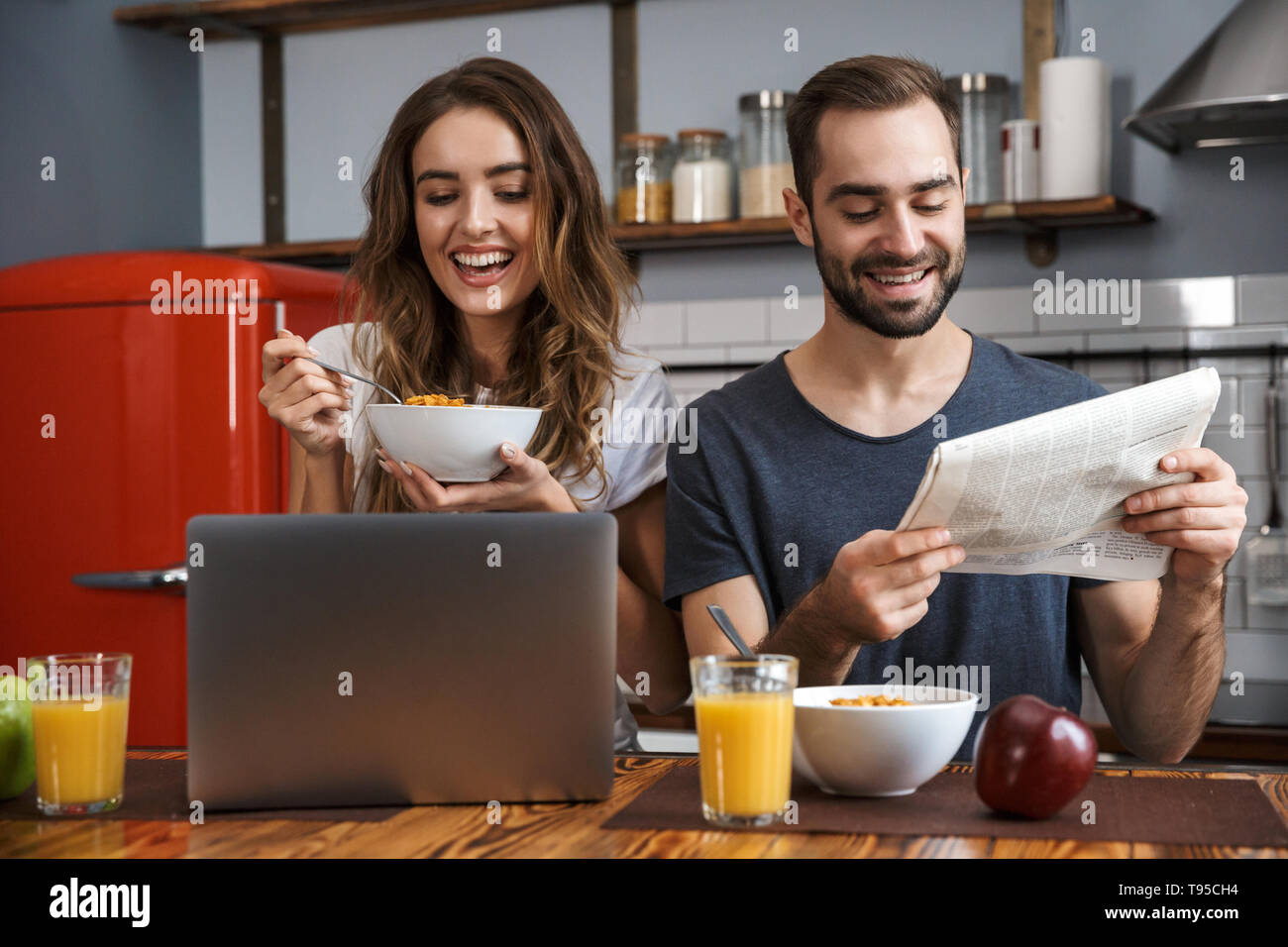 Beautiful cheerful couple having breakfast at the kitchen, using laptop ...