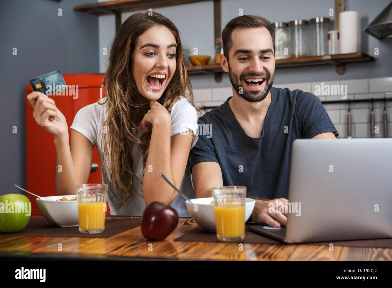 Beautiful cheerful couple having breakfast at the kitchen, using laptop ...