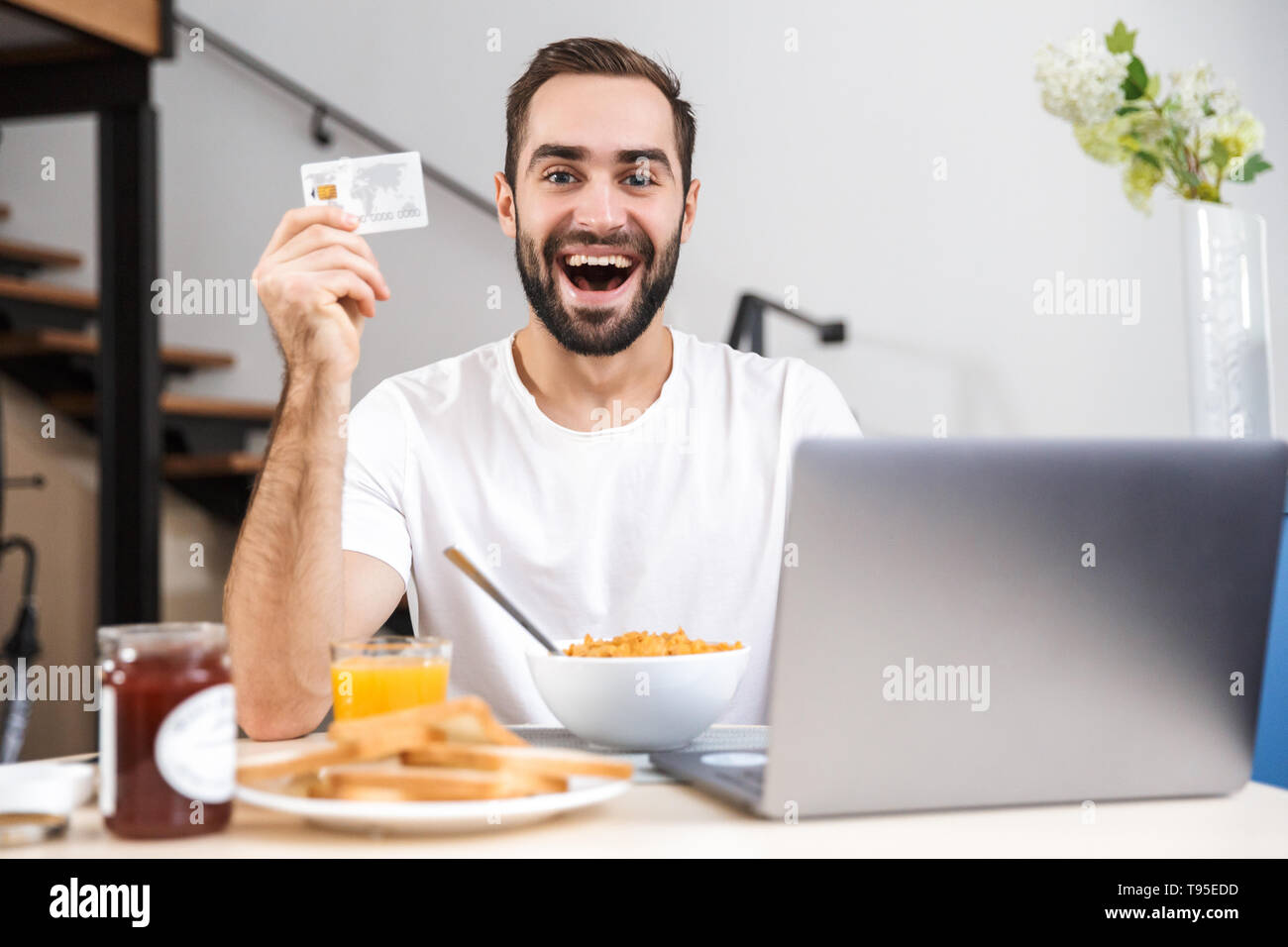 Happy young man having breakfast at the kitchen, using laptop computer ...