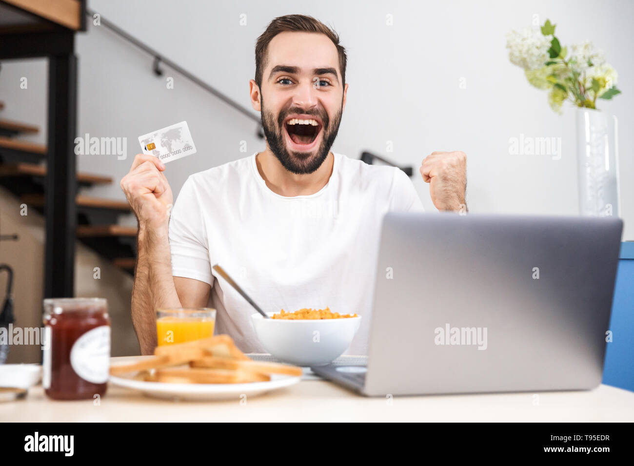 Happy young man having breakfast at the kitchen, using laptop computer ...