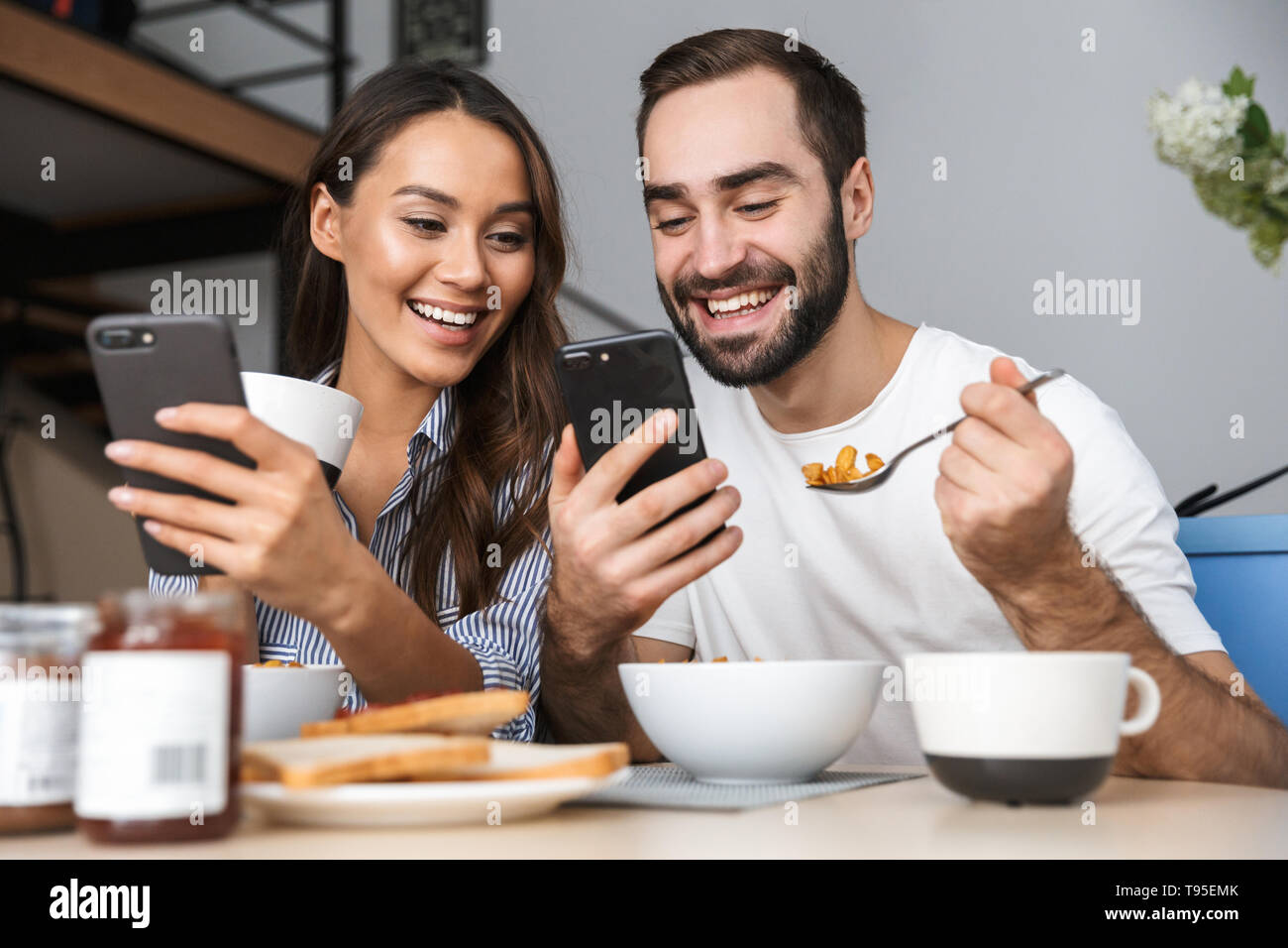 Happy multiethnic couple having breakfast at the kitchen, using mobile ...