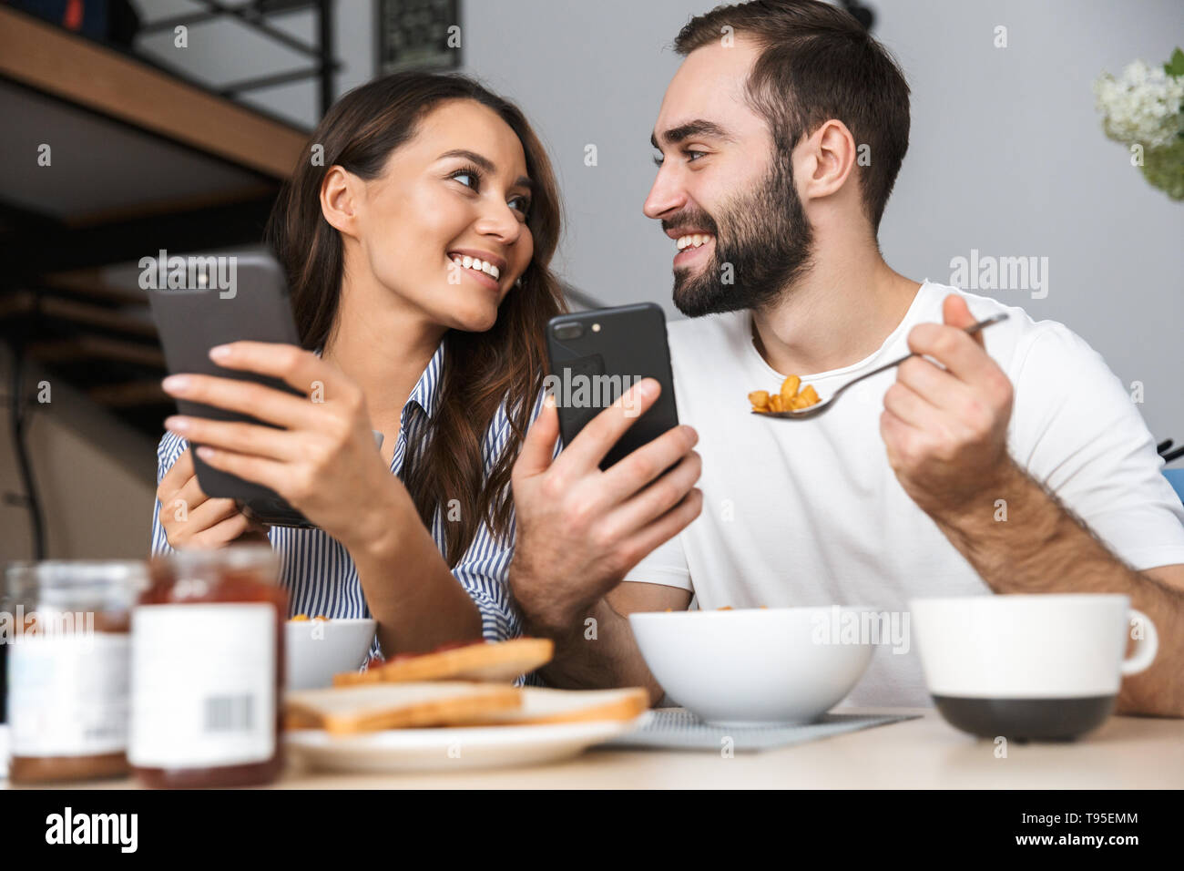 Happy multiethnic couple having breakfast at the kitchen, using mobile ...