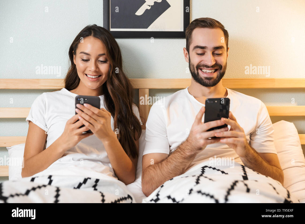 Smiling young couple laying in bed, using mobile phones Stock Photo - Alamy