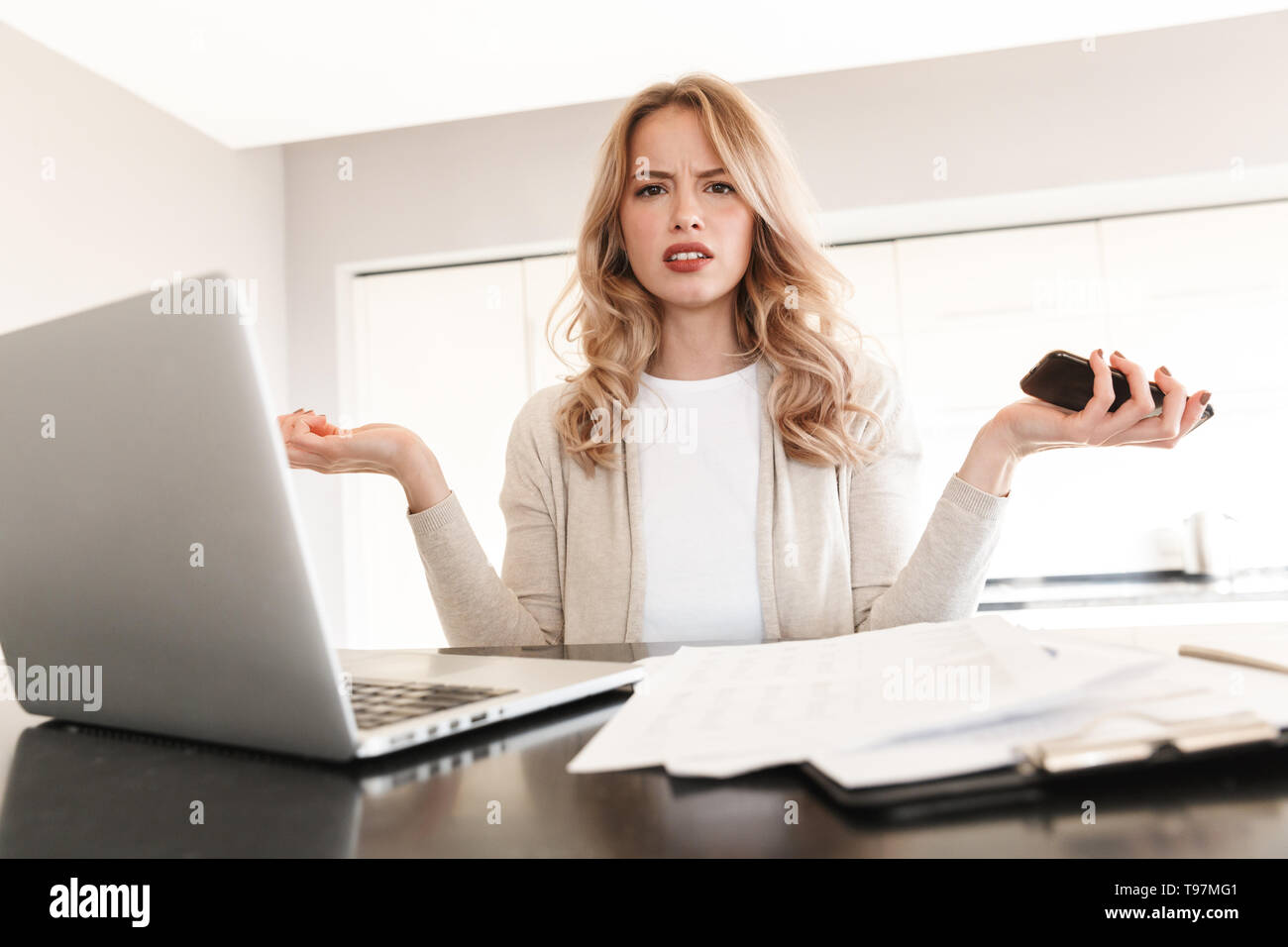 Image of a confused blonde woman posing sitting indoors at home using ...