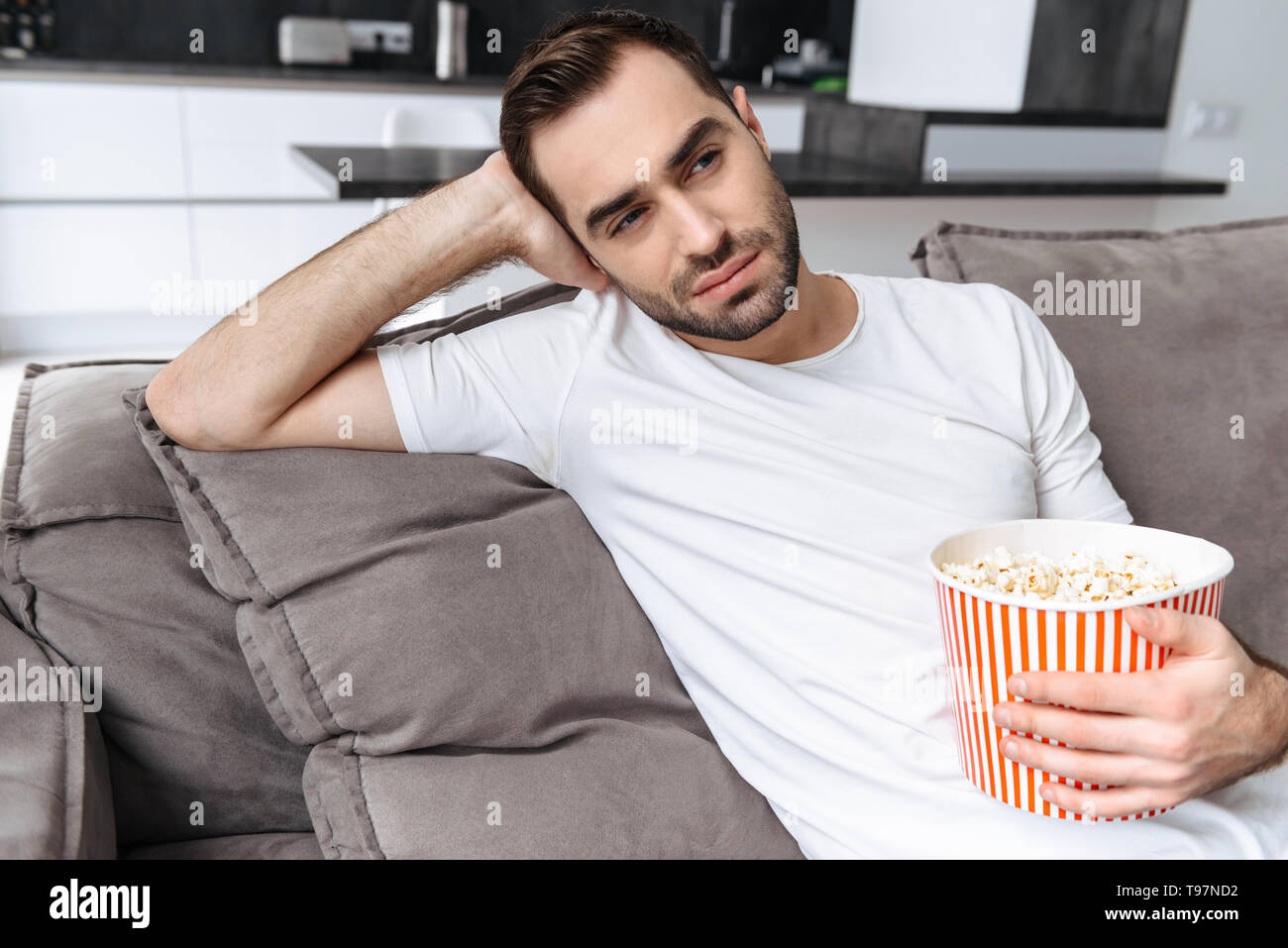 Handsome young man sitting on a couch at home, eating popcorn, watching ...