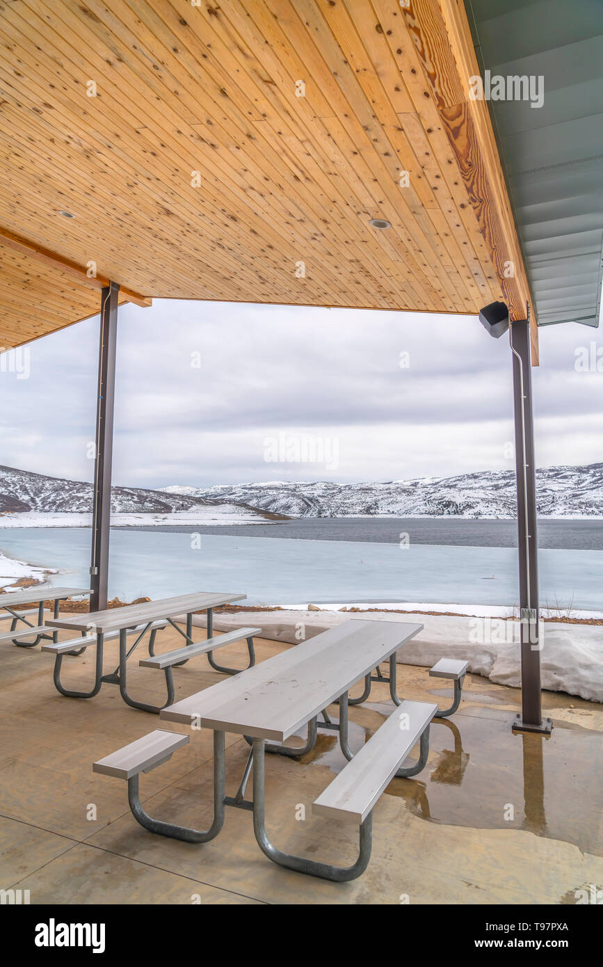 Tables and benches inside a pavilion overlooking an icy lake with snowy ...