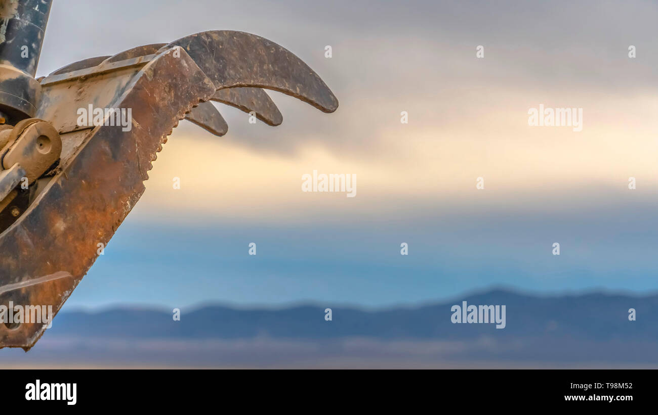 Panorama Excavator bucket teeth against silhouetted mountain and cloudy ...