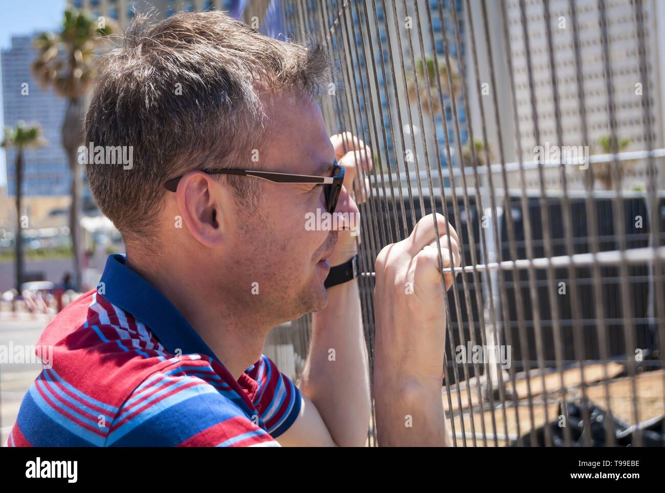 Attractive Caucasian young man looking through the steel grating fence ...