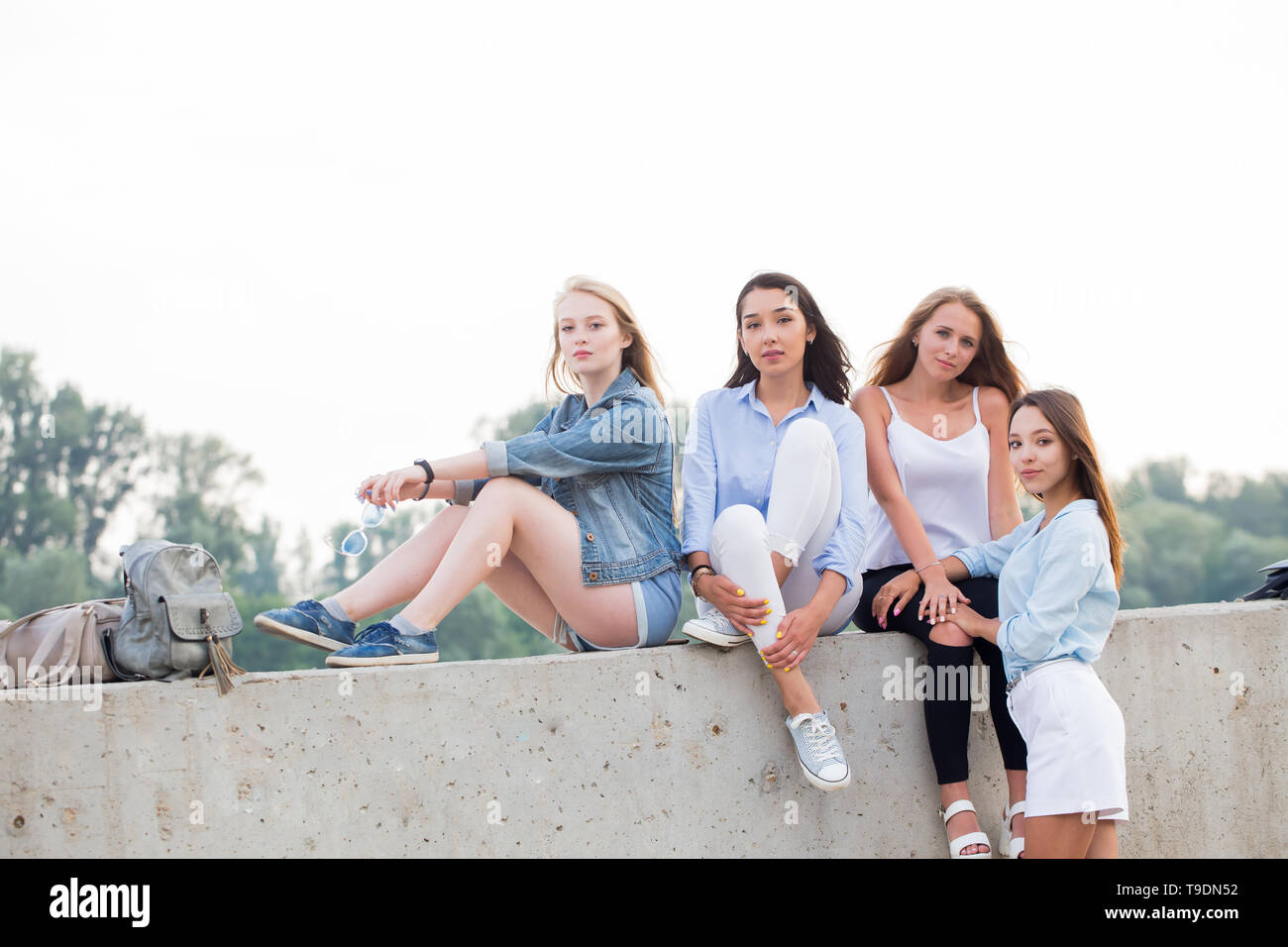 happy cute female friends sitting at concrete border and looking camera ...