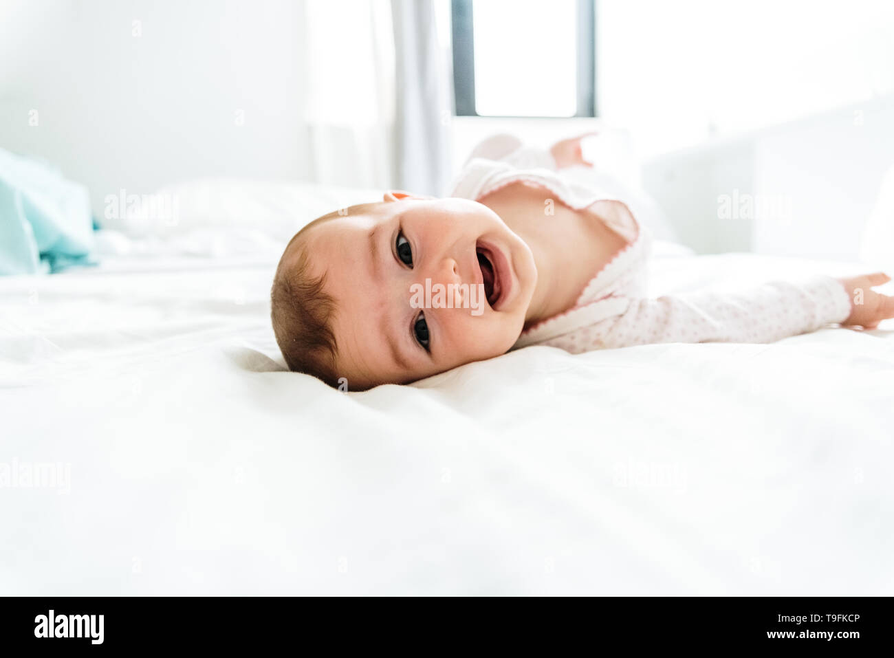 Happy and smiling adorable 6 month old baby girl lying on a bed, lifestyle isolated on natural ...