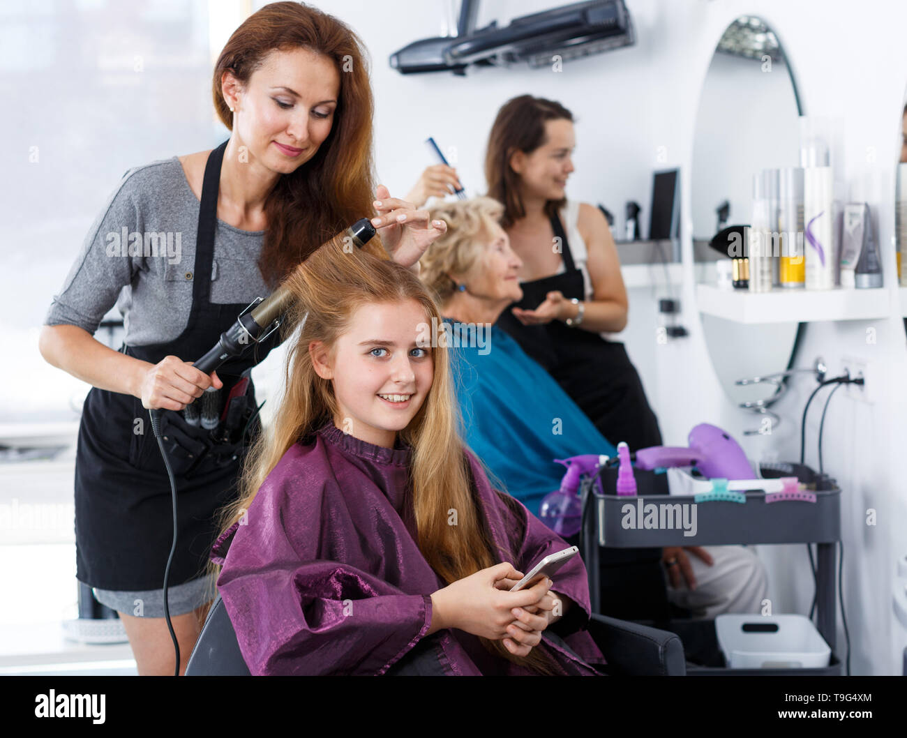 Teenager girl talking on phone while skilled hairdresser curling her ...