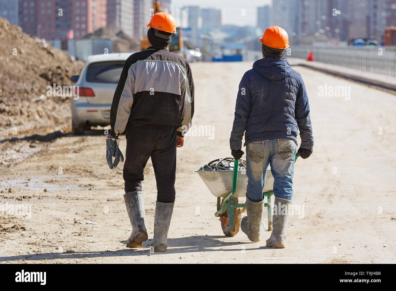 Two workers in helmets drive a wheelbarrow against the background of ...