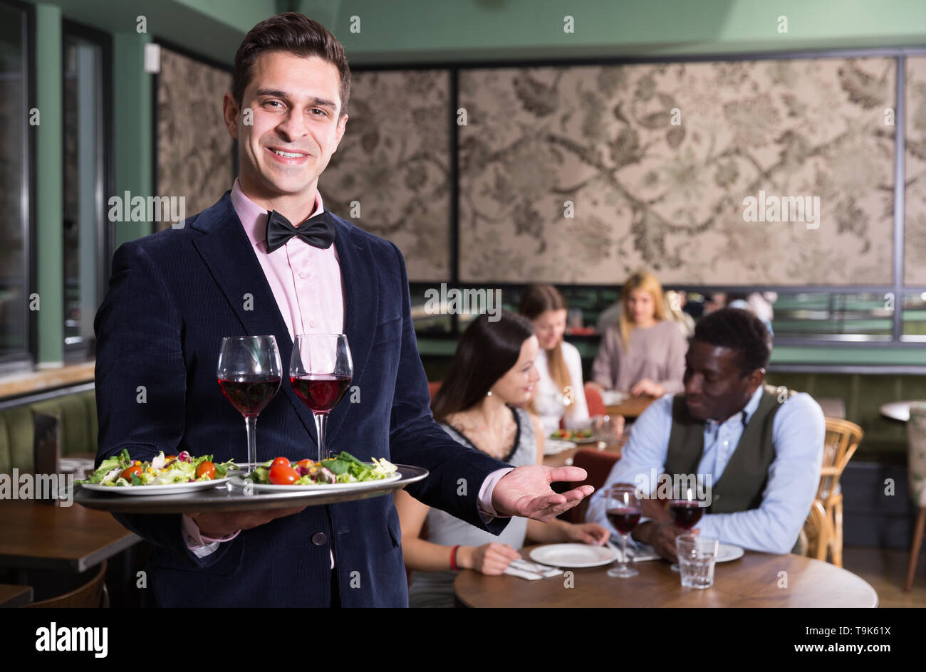 Polite waiter holding tray at restaurant with customers behind him ...