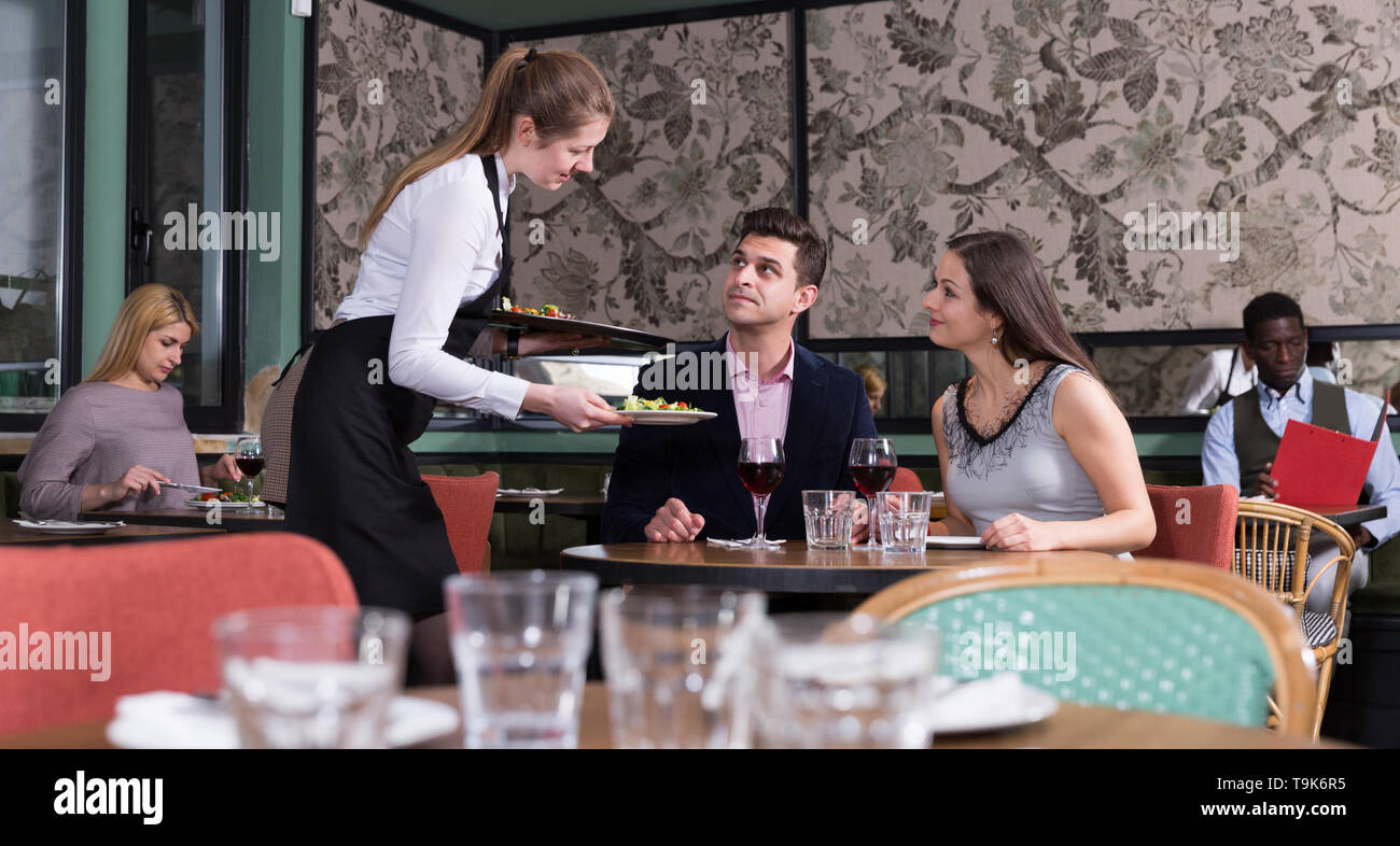Polite waitress bringing ordered dishes to smiling couple at restaurant ...